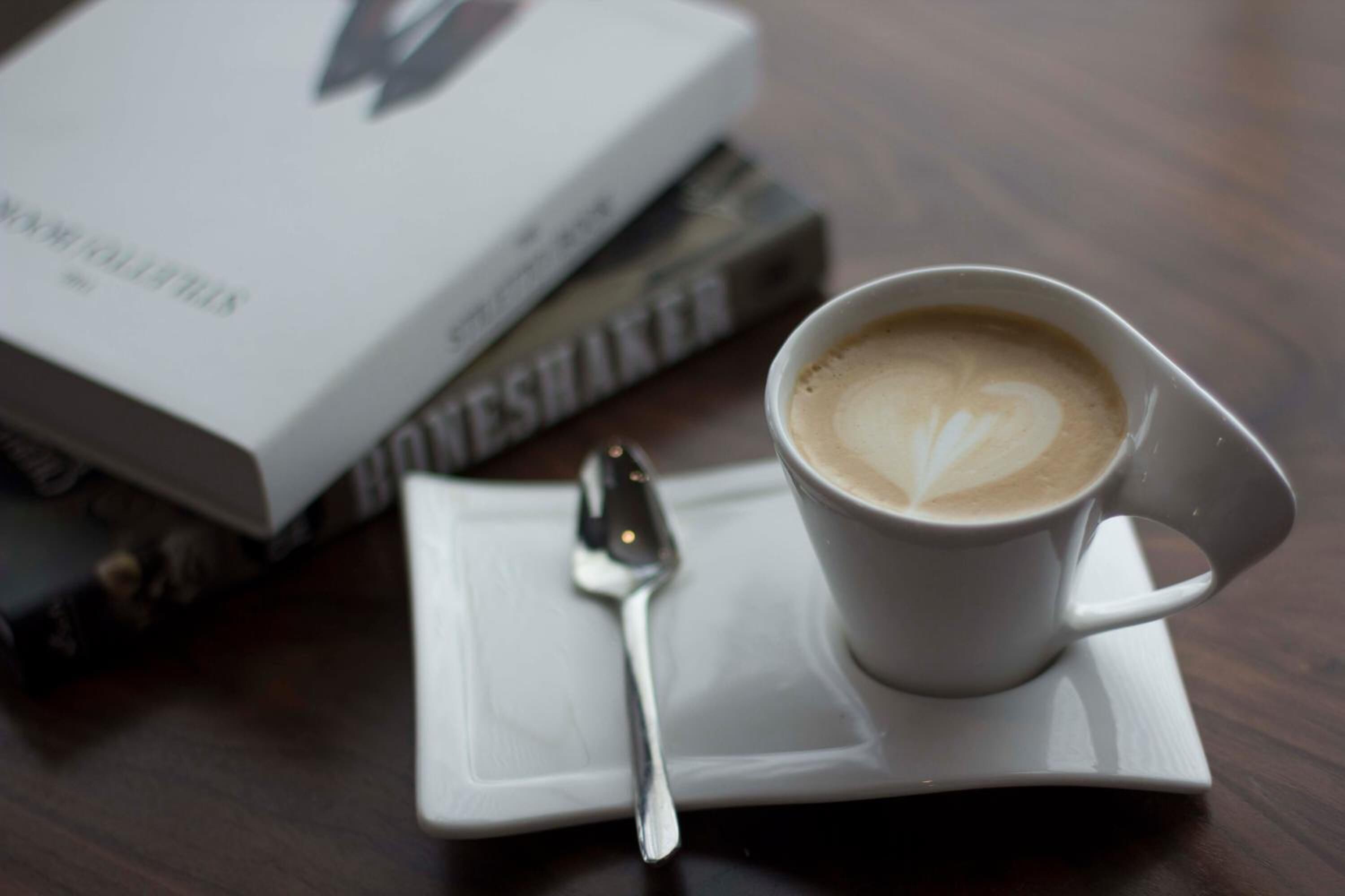 Uma chávena de café com arte de latte num pires, uma colher e uma pilha de livros sobre uma mesa de madeira.