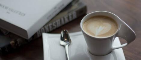 Uma chávena de café com arte de latte num pires, uma colher e uma pilha de livros sobre uma mesa de madeira.