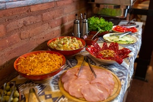 Mesa de buffet com diversos pratos, incluindo uma tigela de queijo ralado, um prato de presunto fatiado e uma tigela de pimentões vermelhos.