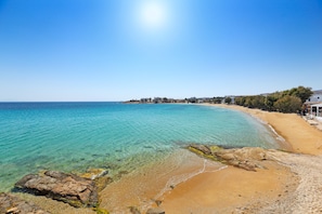 Uma praia com águas turquesa cristalinas, areal arenoso e edifícios ao fundo.