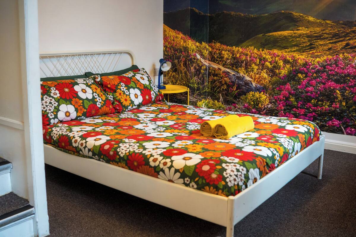 Uma cama com colcha florida, uma toalha amarela e uma mesa de cabeceira com abajur.