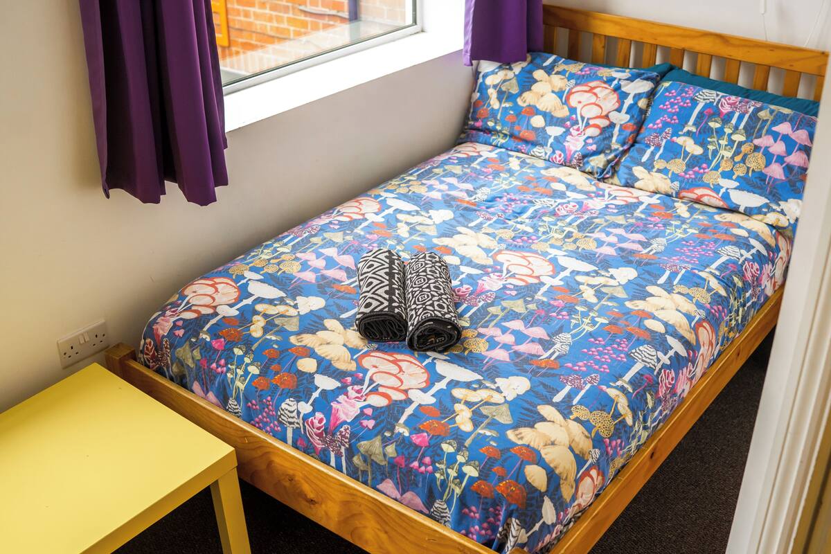 Uma cama de solteiro com colcha florida, cabeceira de madeira e uma pequena mesa amarela ao lado.