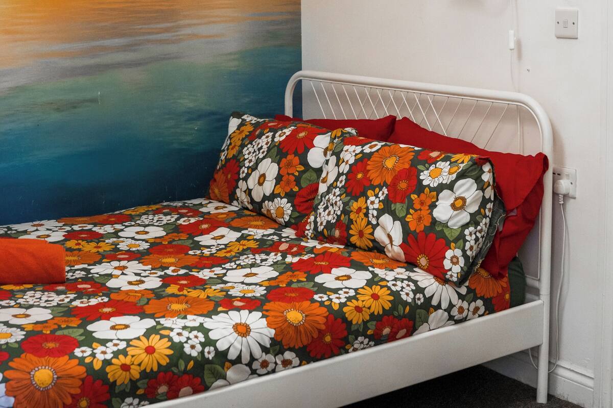Uma cama com colcha florida, estrutura de metal branca, luminária na parede e um mural com um pôr do sol.