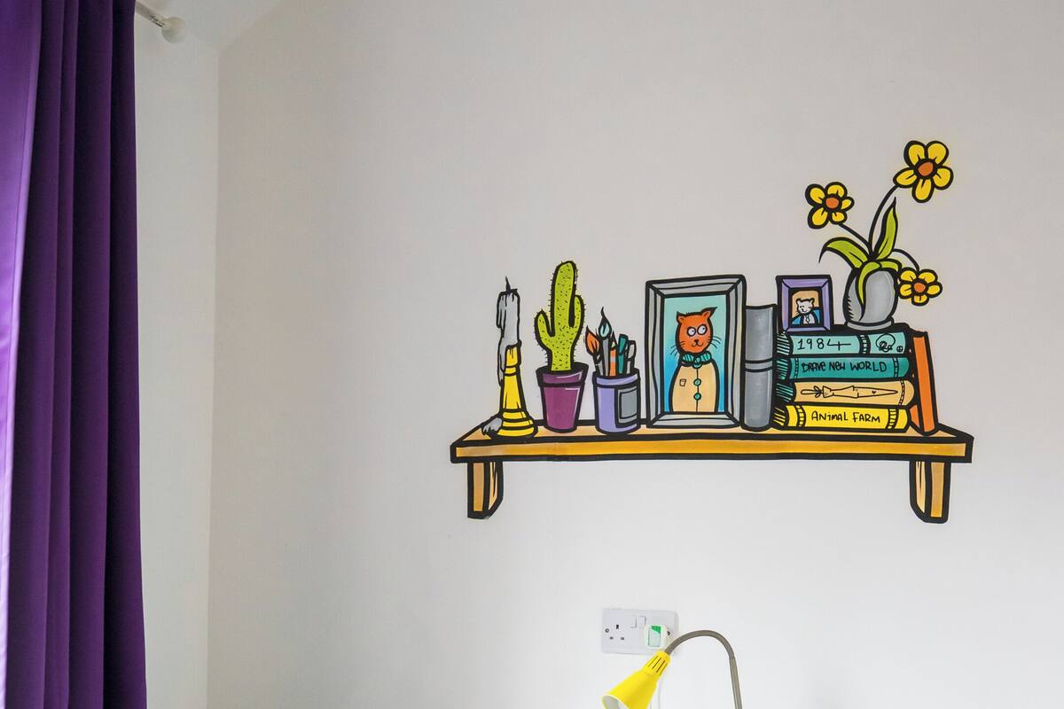 Um quarto com uma escrivaninha branca, duas cadeiras verdes, um abajur amarelo, uma cortina roxa e um ornamento na parede acima da escrivaninha.