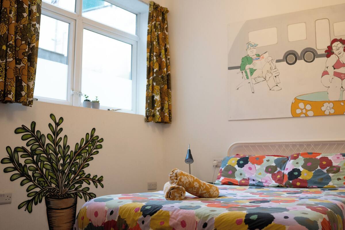 Quarto com cama, planta em vaso, pintura na parede e janela com cortinas.