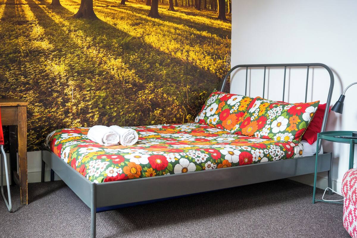 Quarto com cama, cadeira vermelha, escrivaninha e um mural na parede.