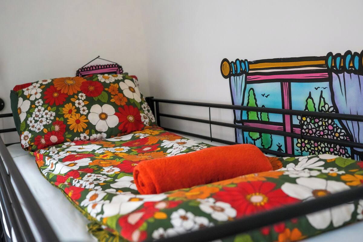 Cama com colcha florida, travesseiro colorido e cabeceira com vista para a janela.