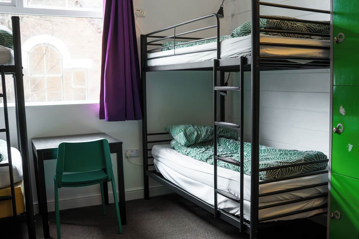 Quarto com beliche, escrivaninha e cadeira, janela com cortinas e um armário verde para guardar objetos.