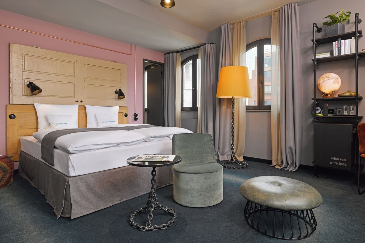 Quarto de hotel com cama, escrivaninha, cadeira, abajur e estante.