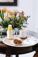 Cenário de café da manhã com um croissant, café, uma garrafa de suco e um arranjo floral sobre uma mesa.