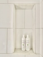 Casa de banho com azulejos brancos e uma prateleira com dois frascos de champô e condicionador Carina Organics.