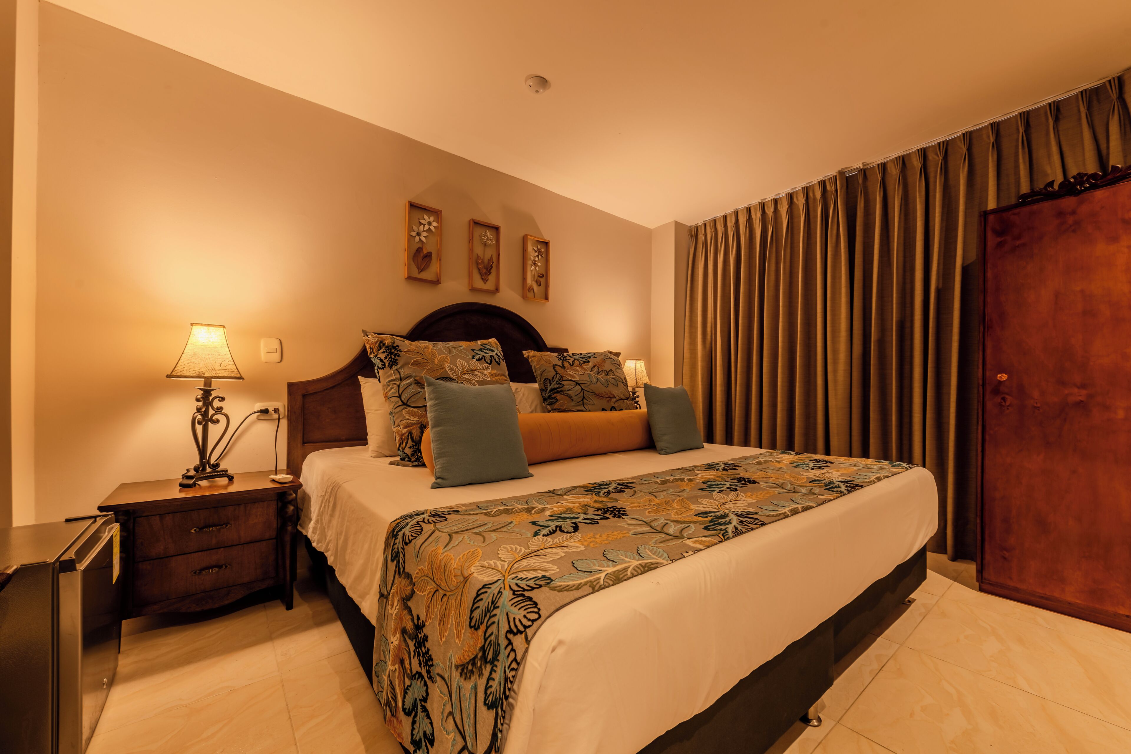 Quarto de hotel com cama, mesa de cabeceira, luminária, guarda-roupa e janela com cortinas.
