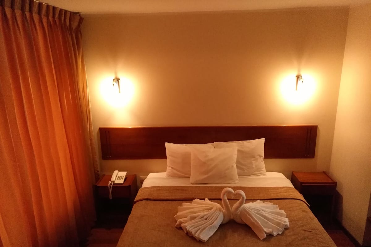 Quarto de hotel com cama bem arrumada, duas luminárias de cabeceira, um telefone e arranjo de toalhas em formato de cisne decorativo.