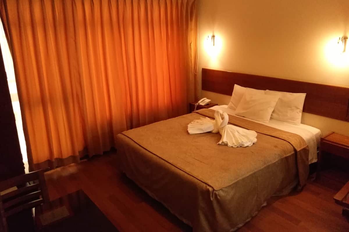 Quarto de hotel com cama, mesa de cabeceira com telefone e janela com cortinas.