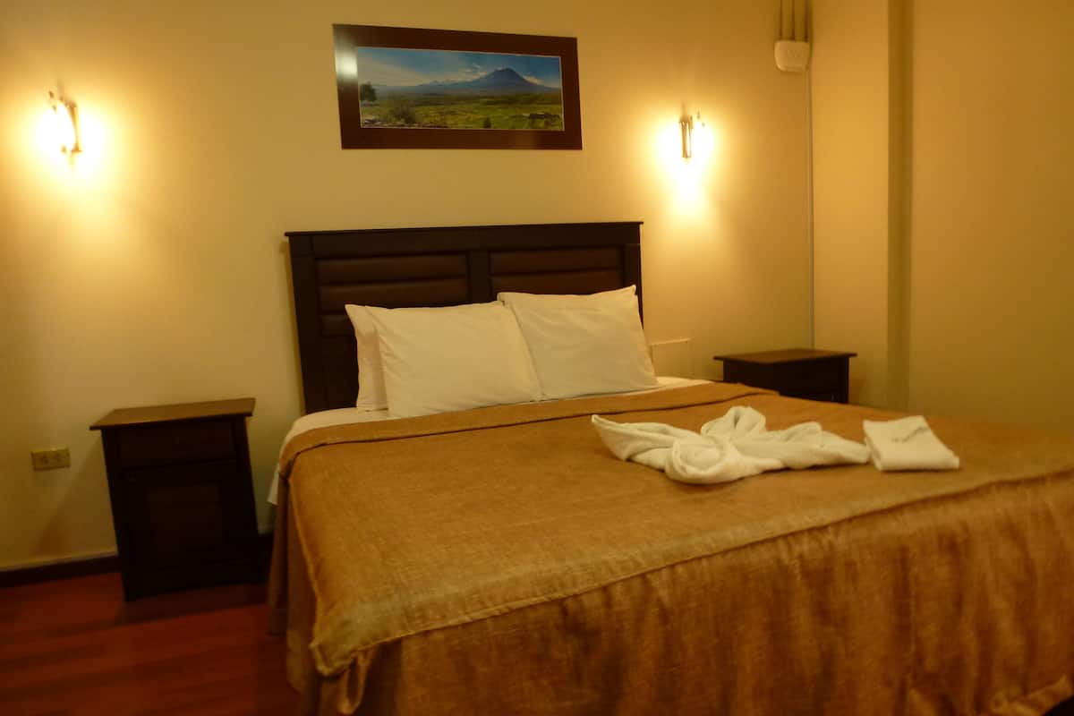 Cama bem arrumada com um cobre-leito marrom, duas almofadas brancas e uma toalha dobrada. Um criado-mudo com uma luminária está à esquerda da cama. Uma moldura com um quadro na parede acima da cama.