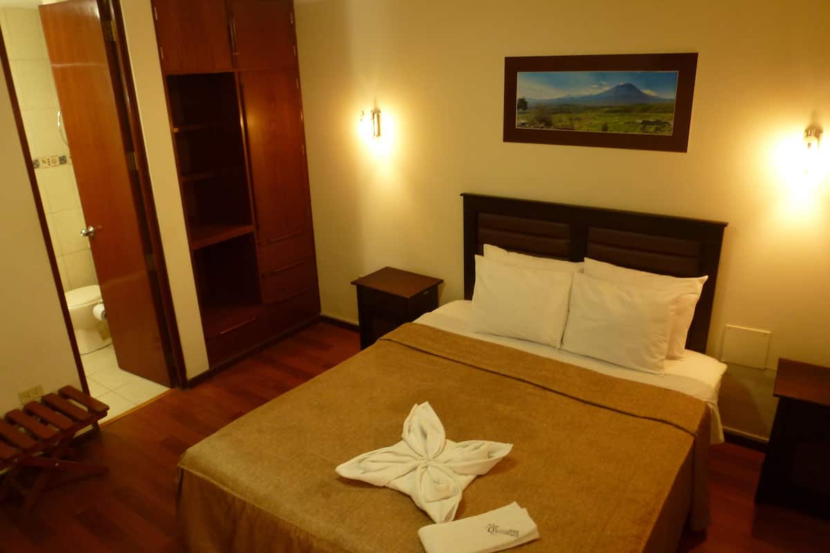 Quarto de hotel com cama, guarda-roupa de madeira, mesa de cabeceira e um quadro na parede.
