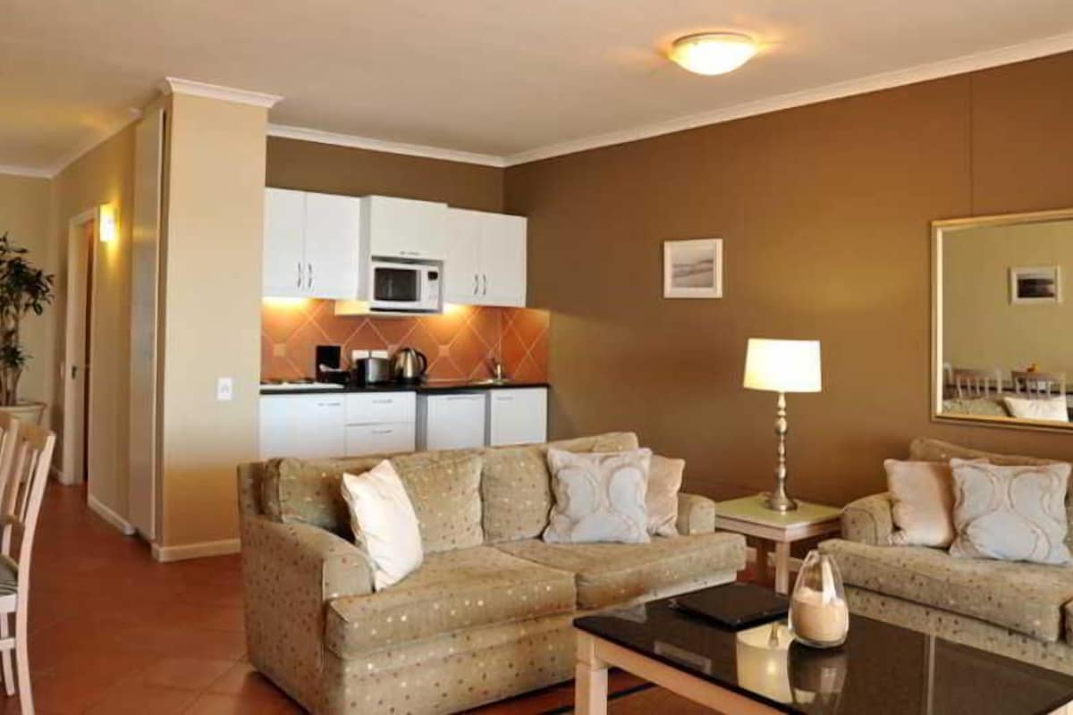 Sala de estar com sofá, mesa de centro e luminária. Há uma área de cozinha com armários e micro-ondas.
