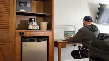 Quarto de hotel com um armário de madeira contendo uma cafeteira, uma chaleira e um micro-ondas. Há também uma geladeira, uma escrivaninha com um laptop e uma cama com uma bolsa preta.
