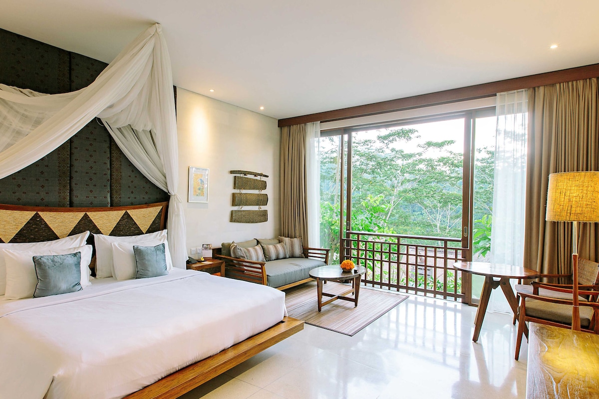 Quarto com cama grande, sofá, varanda e vista para o verde.