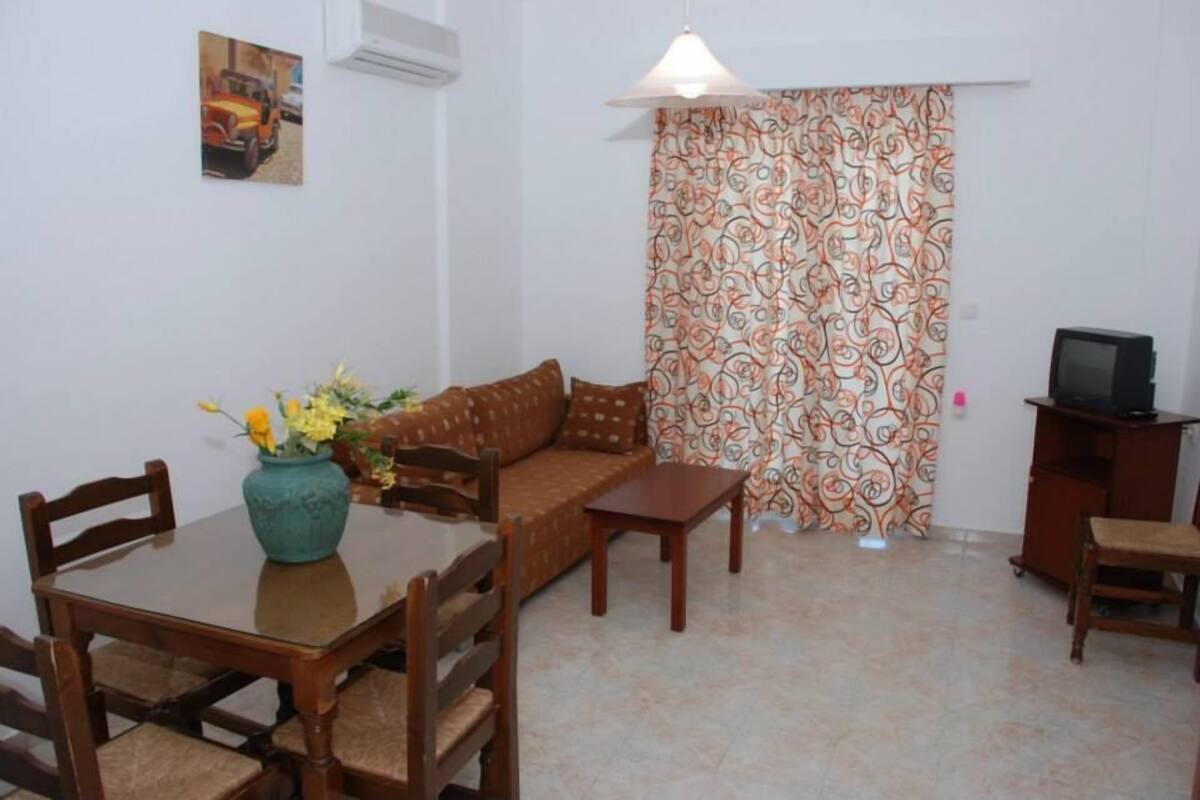 Uma sala de estar com sofá, mesa de jantar com cadeiras, televisão sobre um móvel e cortina estampada.