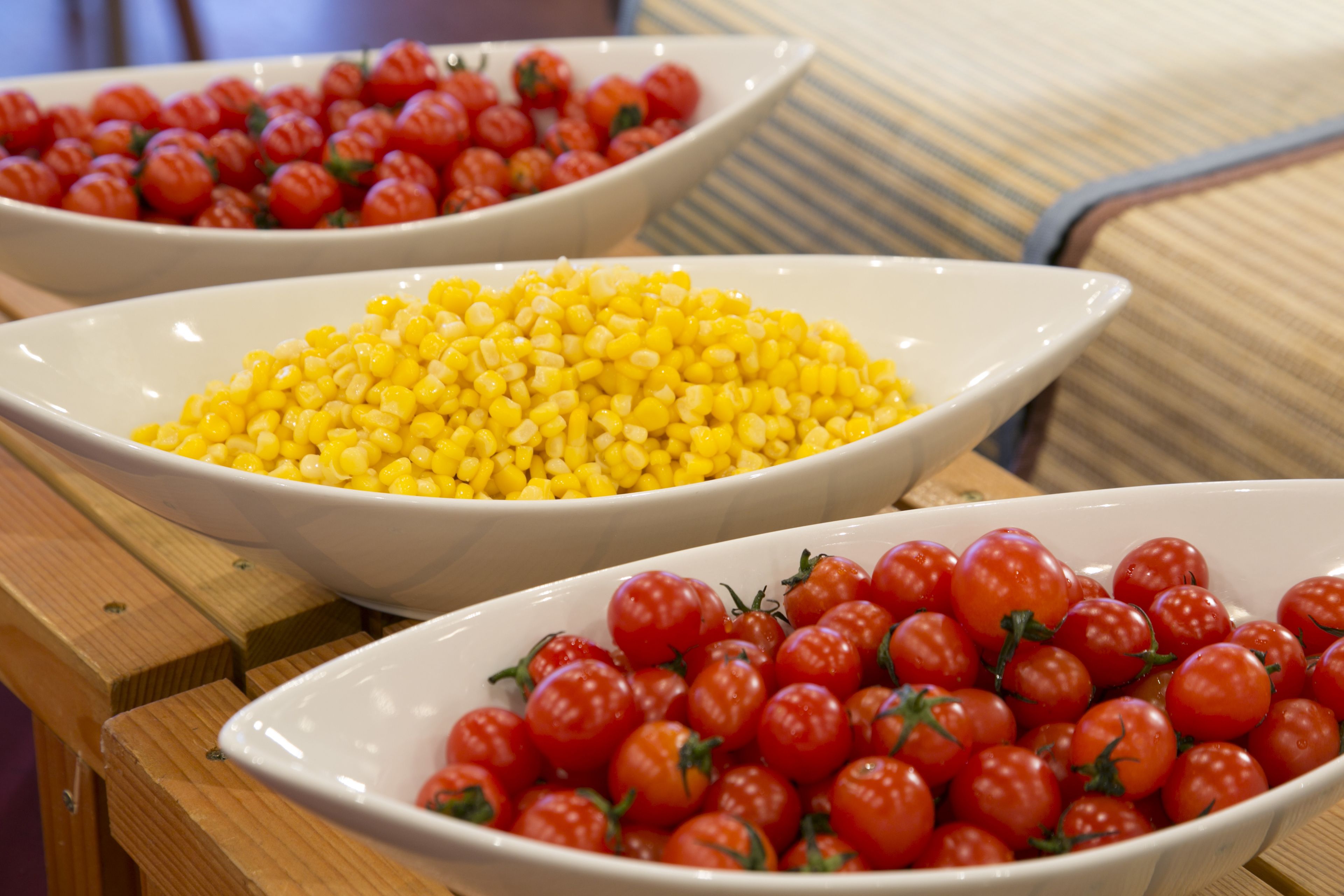 Três tigelas brancas sobre uma mesa de madeira, cada uma contendo itens diferentes: uma com grãos de milho amarelos, outra com tomates cereja vermelhos e a terceira com uma mistura de ambos.
