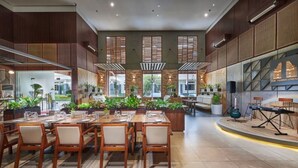 Um restaurante espaçoso com pé-direito alto, painéis de madeira e grandes janelas. As mesas estão dispostas com cadeiras e há plantas e um palco com instrumentos musicais.
