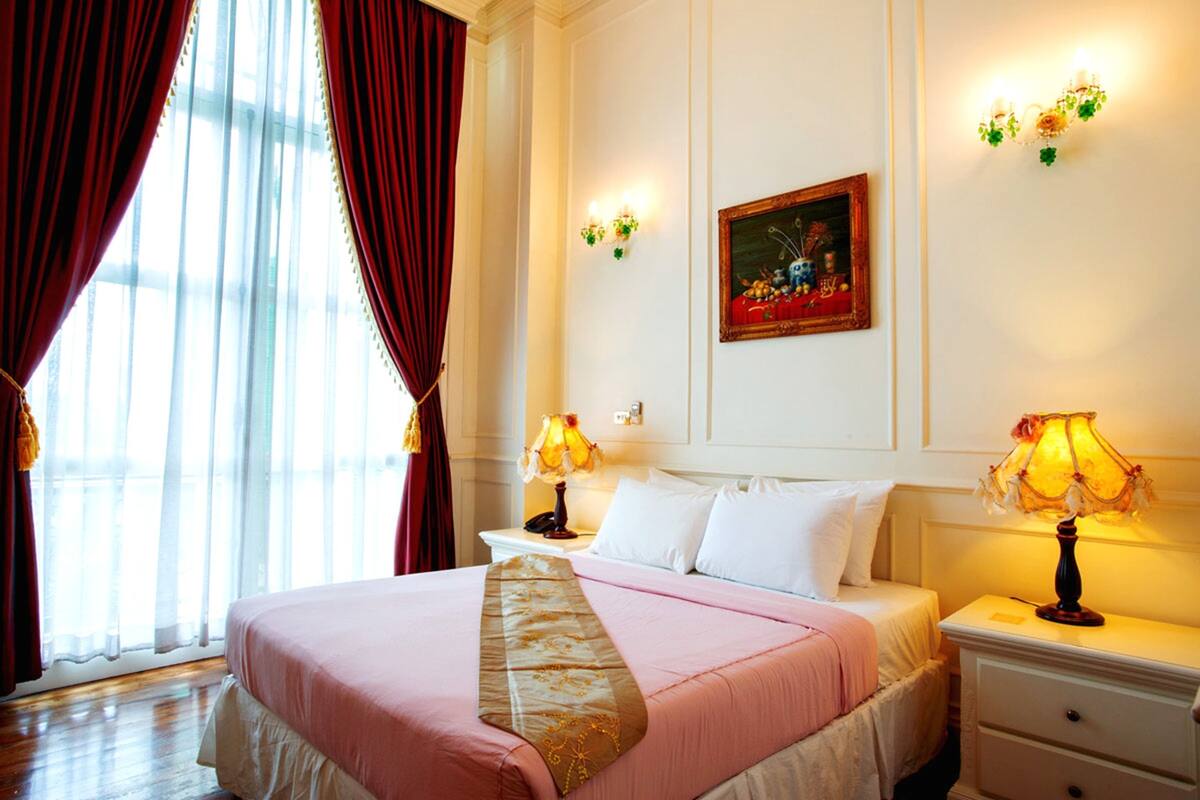 Quarto de hotel com uma cama grande, duas mesas de cabeceira com luminárias, um quadro na parede e uma janela com cortinas.