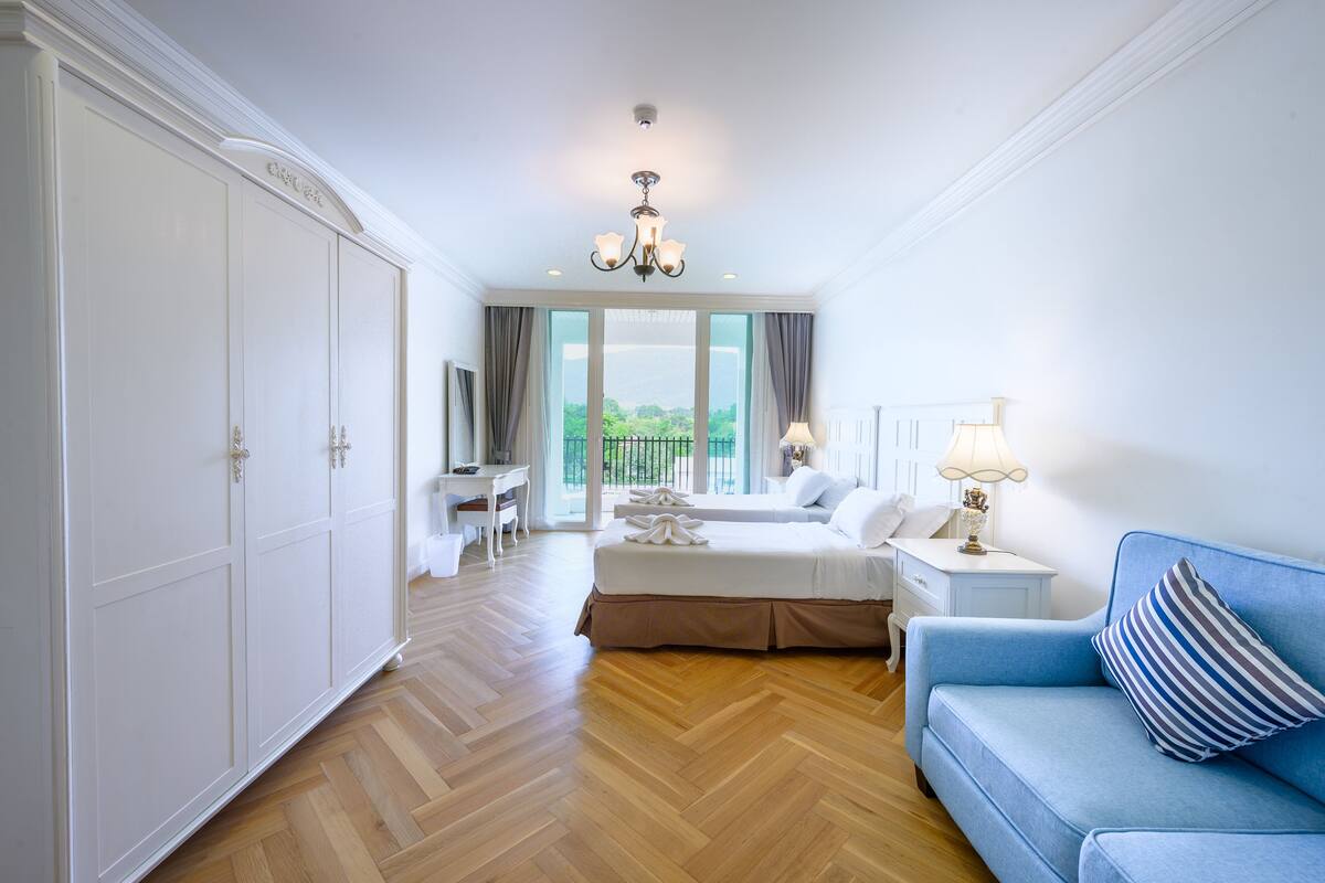 Quarto de hotel com um guarda-roupa espaçoso, uma cama com saia marrom, um sofá azul com almofadas listradas, uma escrivaninha com cadeira e uma janela grande com cortinas.