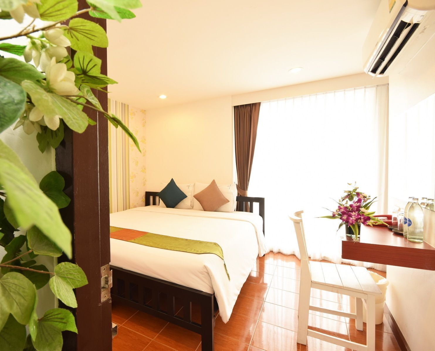 Quarto de hotel com cama, mesa de cabeceira com um vaso de flores, uma cadeira e janela com cortinas.