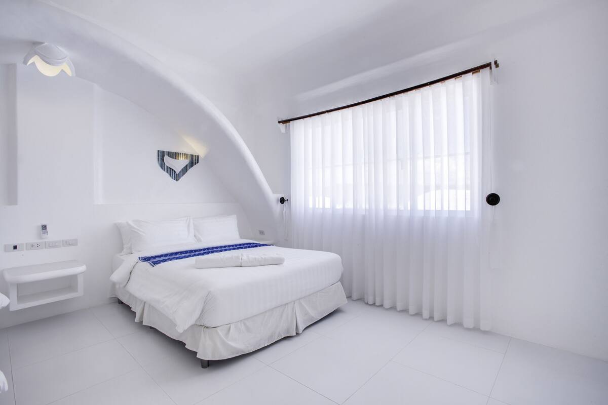 Quarto moderno com uma cama grande, paredes brancas e uma janela com cortinas leves.
