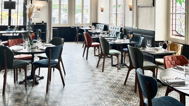Um restaurante com interior clássico, apresentando teto alto com detalhes em gesso ornamentado, grandes janelas com vitrais e diversos tipos de assentos, incluindo mesas e cabines.