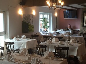 Uma sala de jantar com várias mesas preparadas para o serviço, um quadro grande na parede e uma televisão fixada na parede.