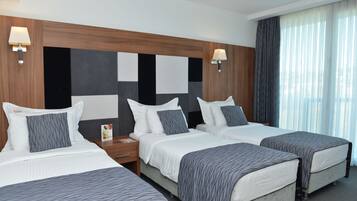 Quarto de hotel com três camas, cada uma com colcha listrada em cinza e branco, cabeceira de madeira e abajures laterais.