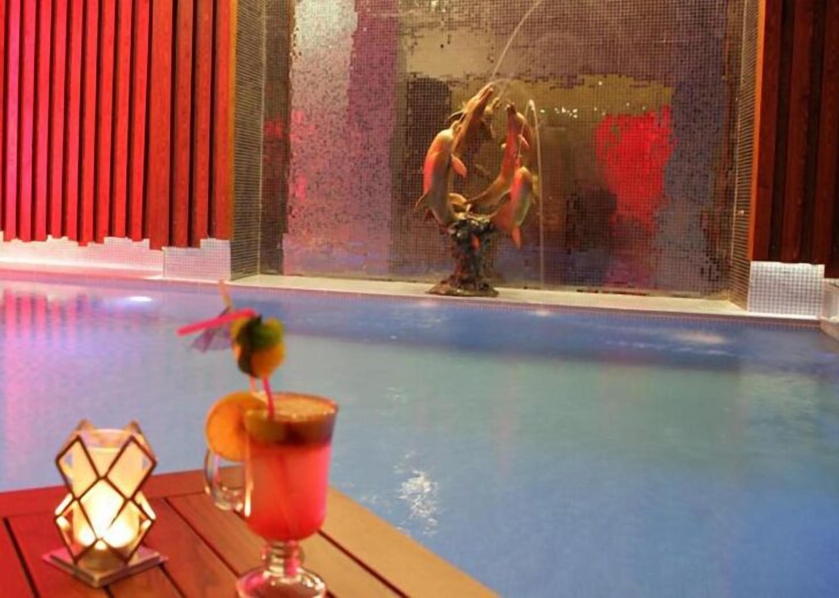 Uma área de piscina com uma estátua decorativa, uma bebida sobre uma mesa e um pano de fundo com cortina vermelha.
