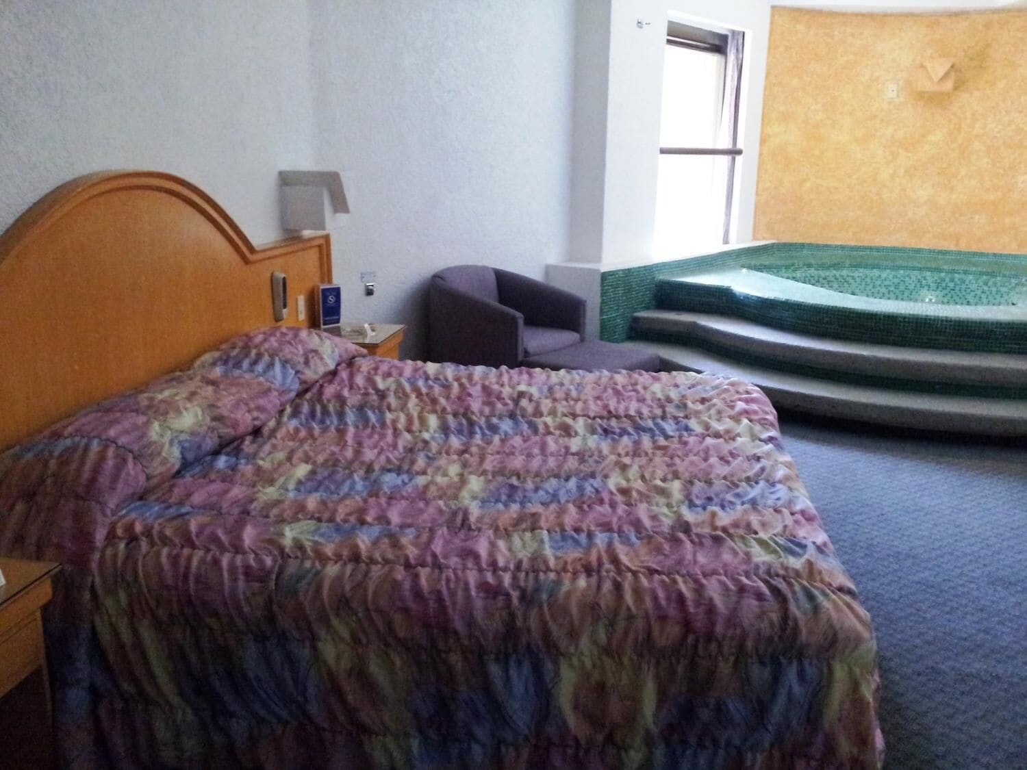 Quarto de hotel com cama de madeira, uma pequena mesa, uma cadeira e área externa verde.