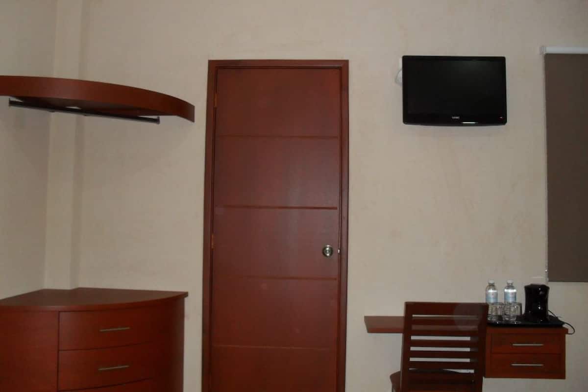 Quarto de hotel com guarda-roupa de madeira, escrivaninha com cadeira, TV de tela plana na parede e pequena mesa com cafeteira e garrafas.