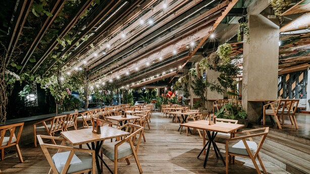Restaurante com mesas e cadeiras de madeira, teto de madeira com luminárias pendentes e plantas em vasos.