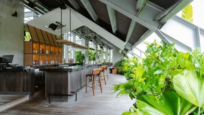 Uma área de bar moderna com banquetas de madeira, teto alto com vigas aparentes e plantas verdes exuberantes.