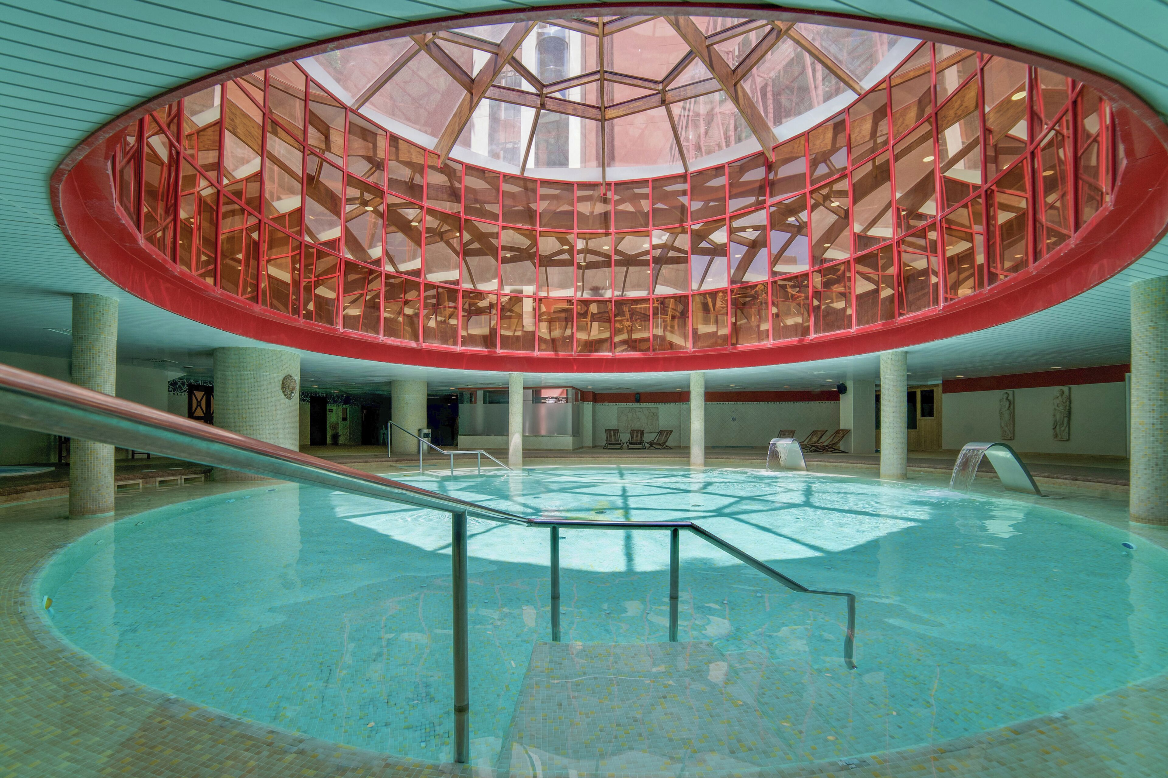 Uma piscina circular com teto de vidro, rodeada por colunas e uma escadaria central.