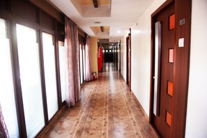 Corredor de hotel com piso de madeira, grandes janelas com cortinas e portas com placas de identificação.