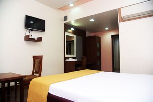 Quarto de hotel com cama, escrivaninha, cadeira, televisão e espelho.