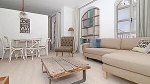 Uma sala de estar com um sofá bege, uma mesa de centro de madeira, cadeiras brancas e uma poltrona estofada. Uma placa na parede diz: "A felicidade não é um destino, é uma forma de viver.".