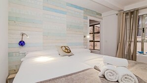 Quarto de hotel com cama, luminária na parede, janela com cortinas e porta-toalhas com toalhas.