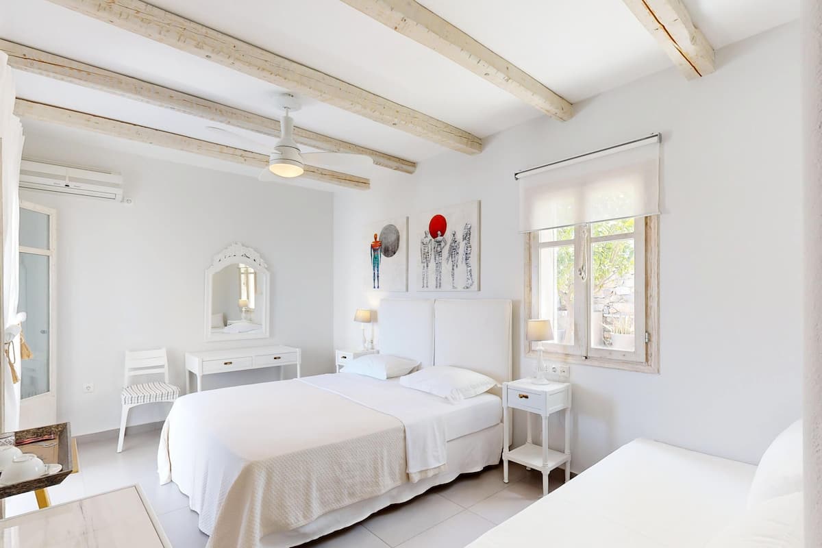 Quarto branco com cama, mesas de cabeceira, cômoda, espelho e janela.