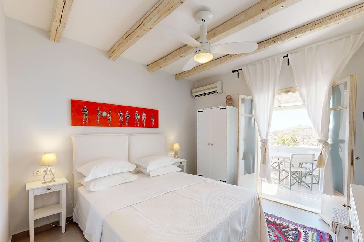 Quarto com cama, mesas de cabeceira brancas, ventoinha no teto, obra de arte vermelha, guarda-roupa e vista pela janela com cortinas.