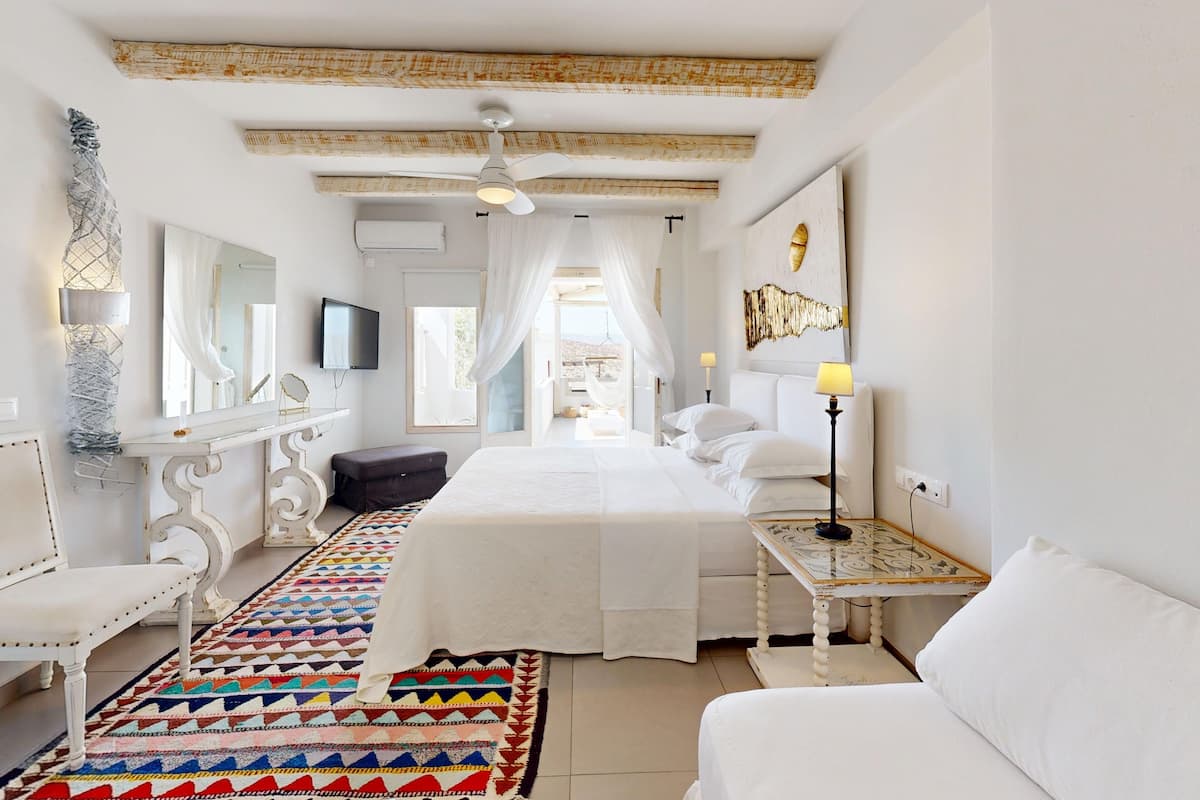 Um quarto espaçoso com uma cama grande, uma mesinha de cabeceira, uma cadeira, um tapete estampado, um ventilador de teto e vista para o exterior.