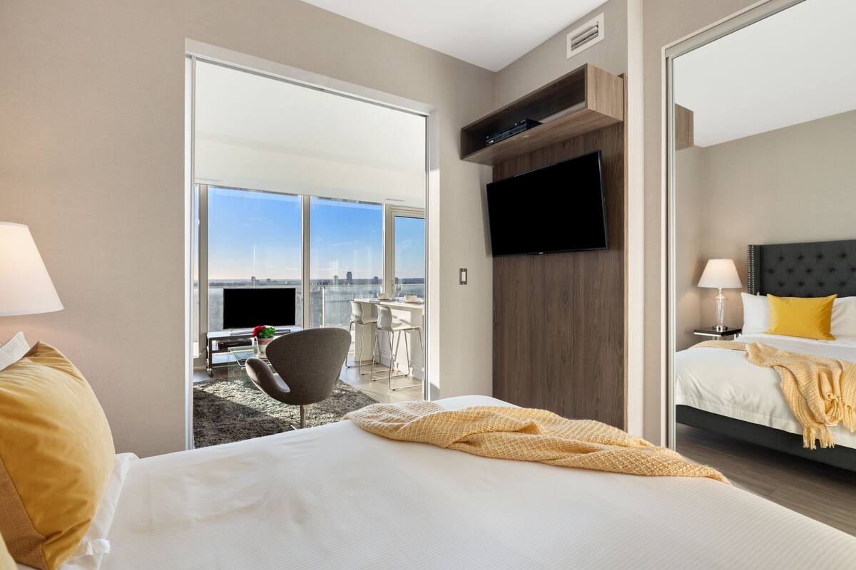 Quarto de hotel com uma cama grande, televisão na parede, escrivaninha com cadeira e vista para a cidade através de grandes janelas.