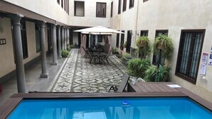 Piscina com deck azulejado, área de varanda coberta com mesas e cadeiras, e plantas em vasos.