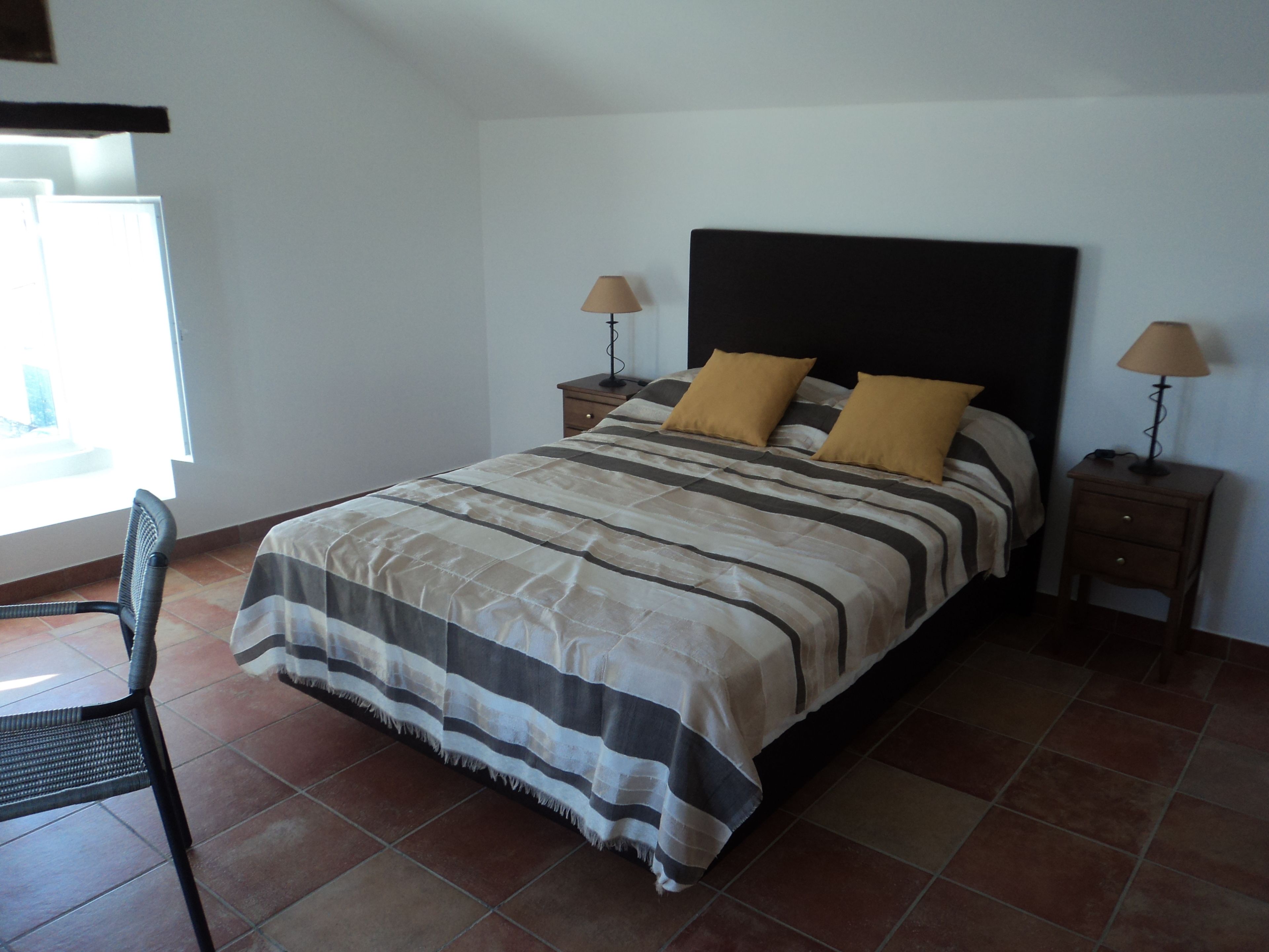 Um quarto com uma cama, duas mesas de cabeceira, uma cadeira e uma janela.
