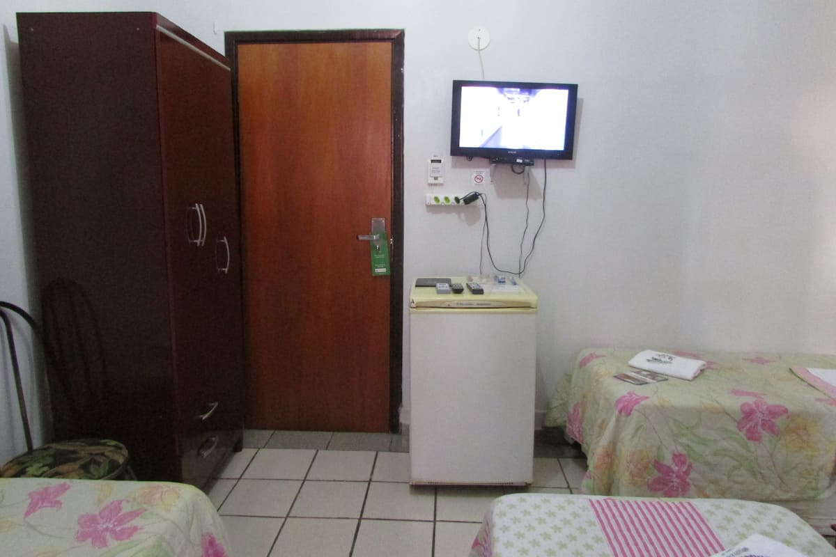 Quarto de hotel com guarda-roupa de madeira, pequeno frigorífico, televisão fixada na parede e duas camas com roupa de cama floral.